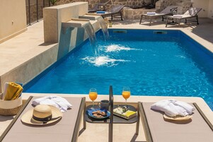 Área da piscina com um elemento aquático, espreguiçadeiras e uma mesa com bebidas e petiscos.