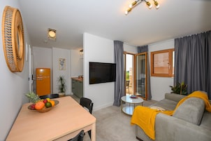 Uma sala de estar moderna com televisão de tela plana, uma área de jantar com uma tigela de frutas e um sofá com uma manta amarela.