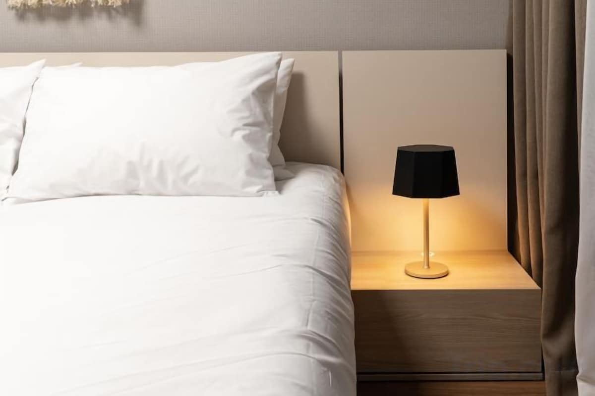 Uma cama bem arrumada com lençóis brancos, uma mesa de cabeceira com abajur preto e um criado-mudo de madeira.