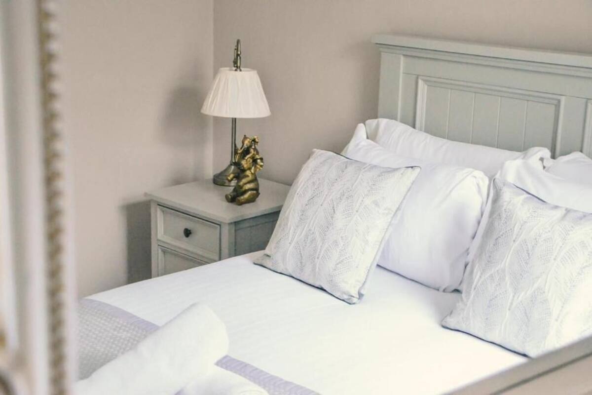 Uma cama bem arrumada com roupa de cama branca e uma luminária decorativa em uma mesinha de cabeceira.