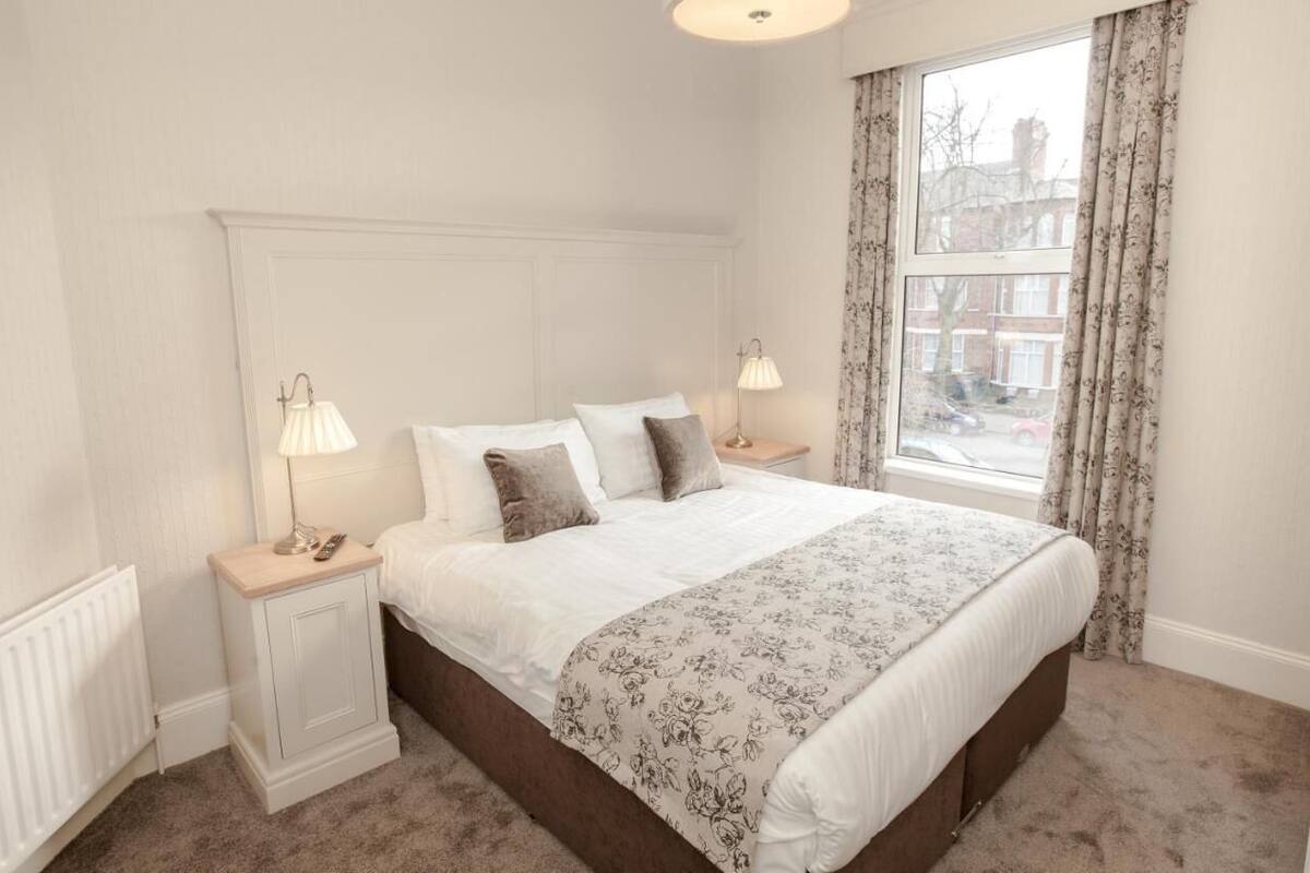 Quarto com cama grande, duas mesas de cabeceira, janela com cortinas e radiador.