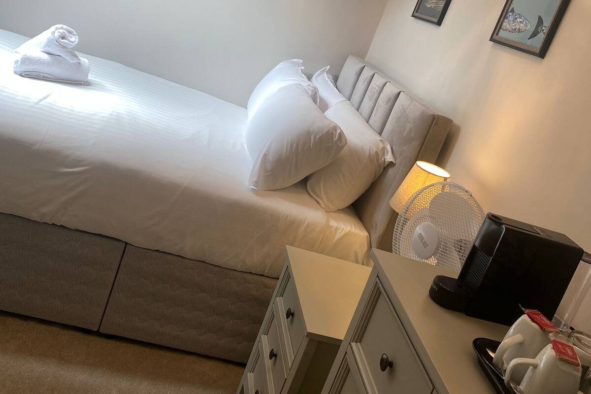 Quarto de hotel com cama, almofadas, mesa de cabeceira com abajur, ventoinha e bandeja com xícaras e um copo.