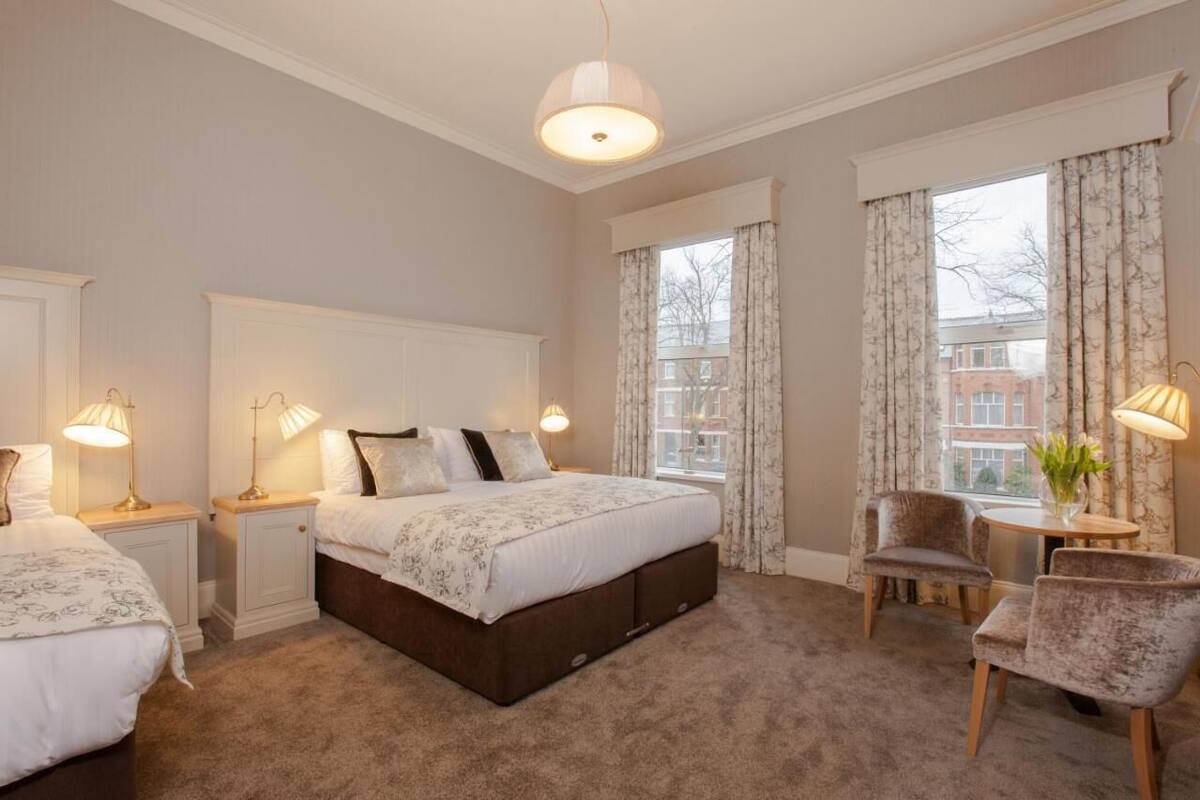 Quarto com uma cama grande, duas mesas de cabeceira com luminárias, uma cadeira, uma mesinha com um vaso e uma janela com cortinas.