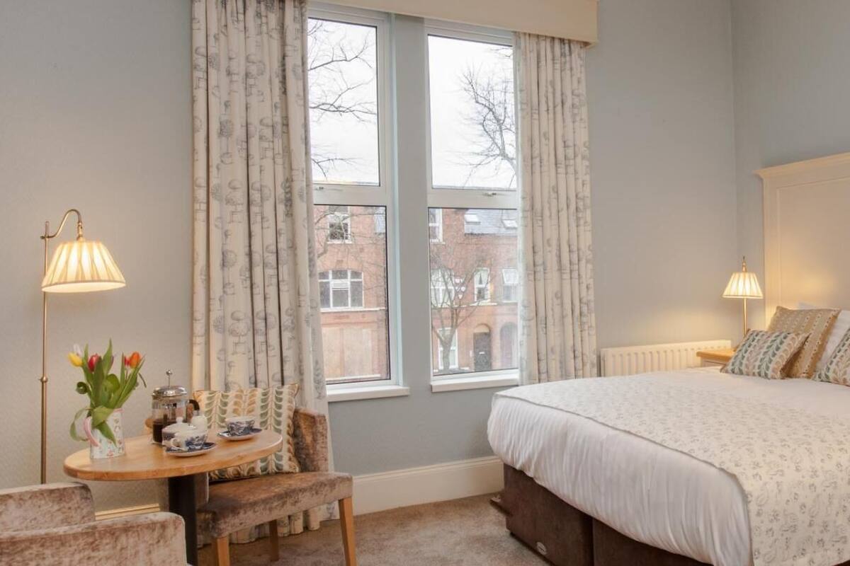 Quarto com uma cama grande, duas luminárias de cabeceira, uma mesa pequena com um vaso de flores e vista para edifícios pela janela.