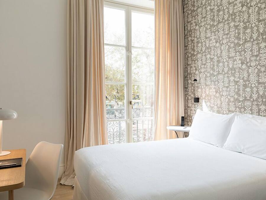 Quarto de hotel com uma cama grande, uma escrivaninha com abajur e vista para o exterior através de uma janela com cortinas.