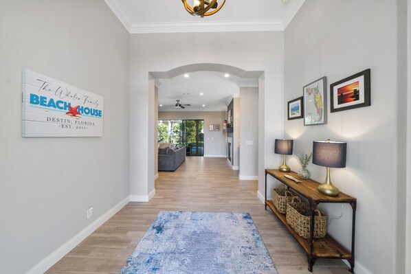 Um corredor com piso de madeira, tapete azul, uma placa com a inscrição "The Whalen Family Beach House Destin, Florida - Est. 2011" e um console com luminárias e cestos.