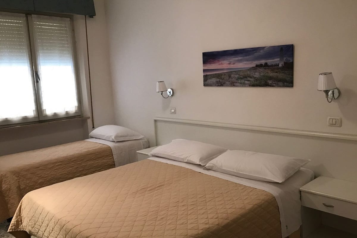 Quarto de hotel com duas camas de solteiro, uma mesa de cabeceira e uma luminária fixada na parede.