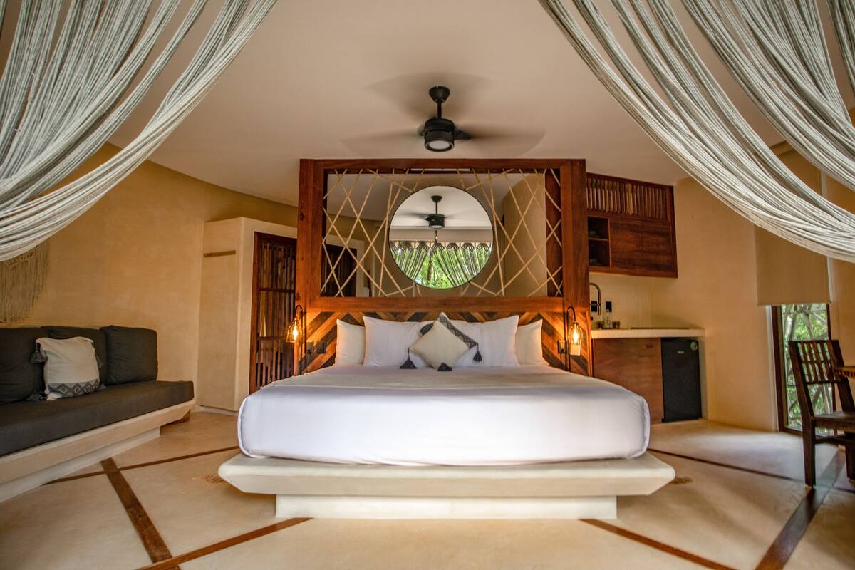 Uma cama grande com cabeceira de madeira, um ventilador de teto e vista para o verde por uma janela circular.