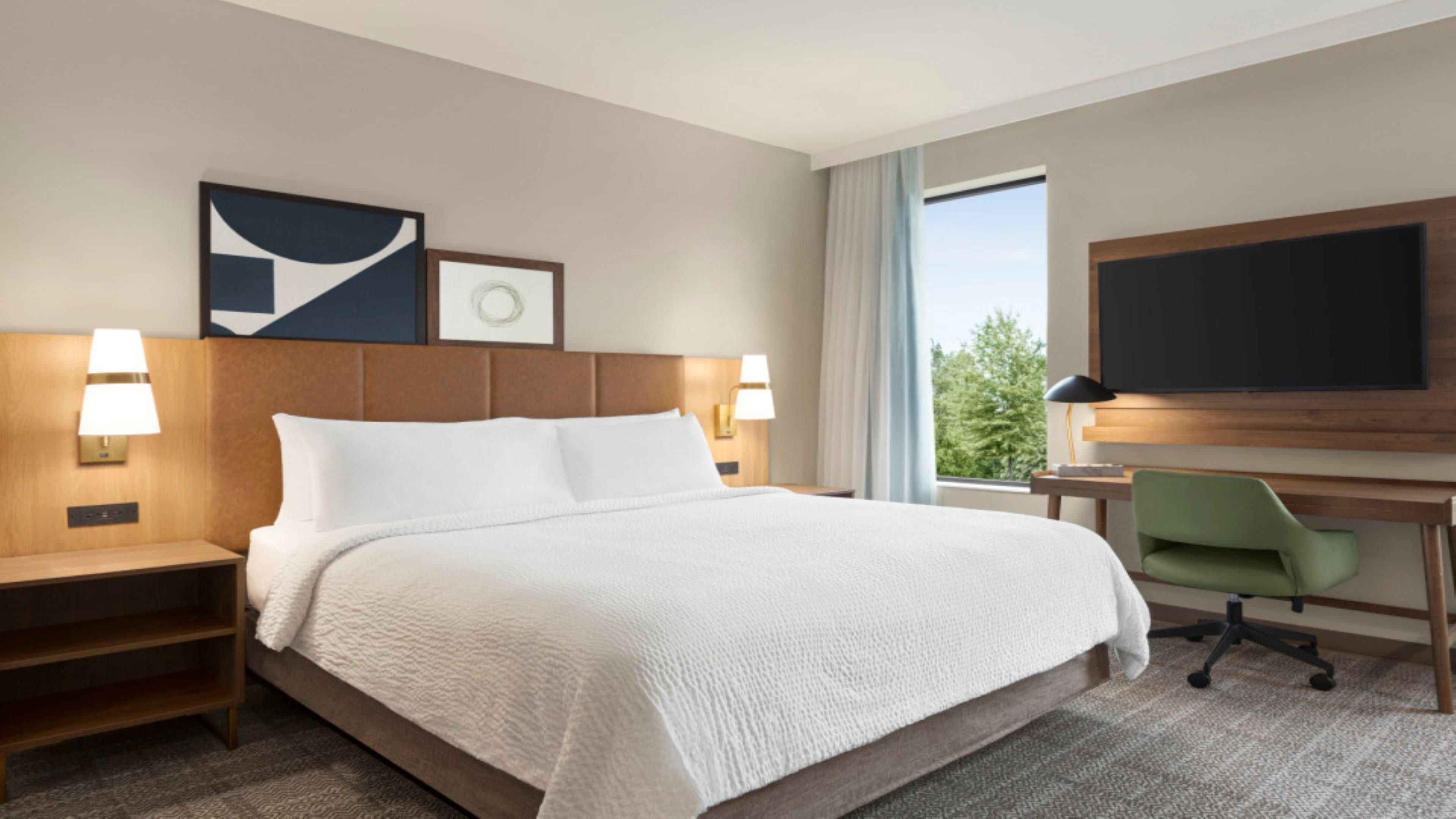 Quarto de hotel com uma cama grande, duas mesas de cabeceira com luminárias, uma escrivaninha com cadeira, uma TV de tela plana e uma janela com vista para árvores.