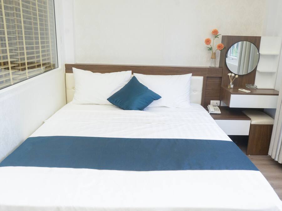Cama bem arrumada com roupa de cama branca e azul, cabeceira de madeira, mesa de cabeceira com espelho e um pequeno vaso com flores.