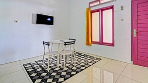 Quarto com piso de azulejos, uma pequena mesa com cadeiras, uma TV de tela plana fixada na parede e uma porta com moldura rosa.