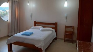 Quarto com cama de madeira, roupa de cama branca, duas luzes fixadas na parede, janela com cortinas e porta de madeira.