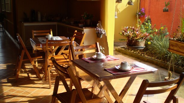 Uma sala de jantar com cadeiras e mesas de madeira, uma cozinha ao fundo e plantas em vasos numa varanda.