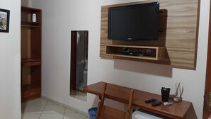 Quarto com um suporte de TV de madeira, uma escrivaninha com computador e uma pequena lixeira.