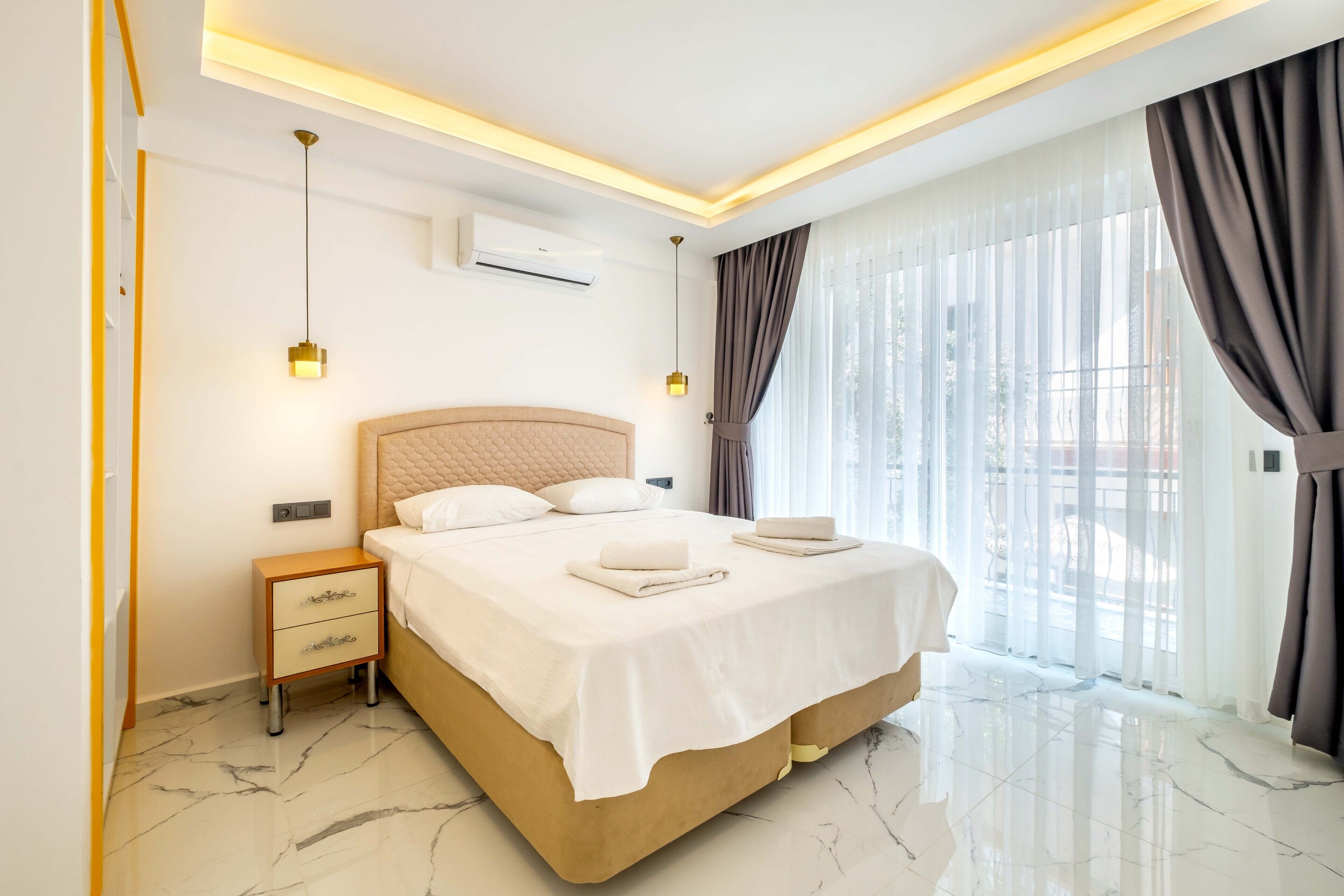Quarto moderno com uma cama grande, mesas de cabeceira, ar condicionado e vista para o exterior através de cortinas leves.