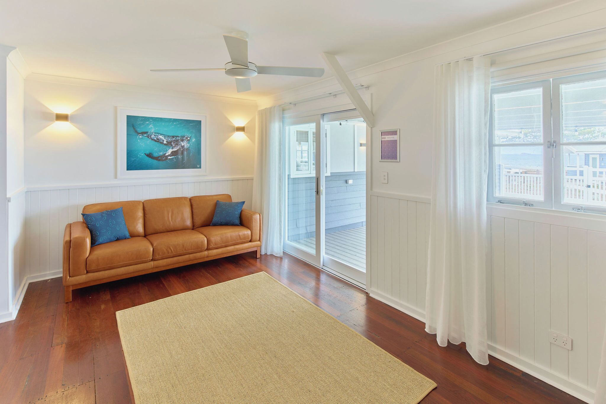 Sala de estar com sofá de couro marrom, tapete bege, ventilador de teto e uma grande janela com cortinas brancas.