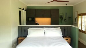 Quarto com cama, mesas de cabeceira de madeira, ventoinha no teto e vista para a cozinha.