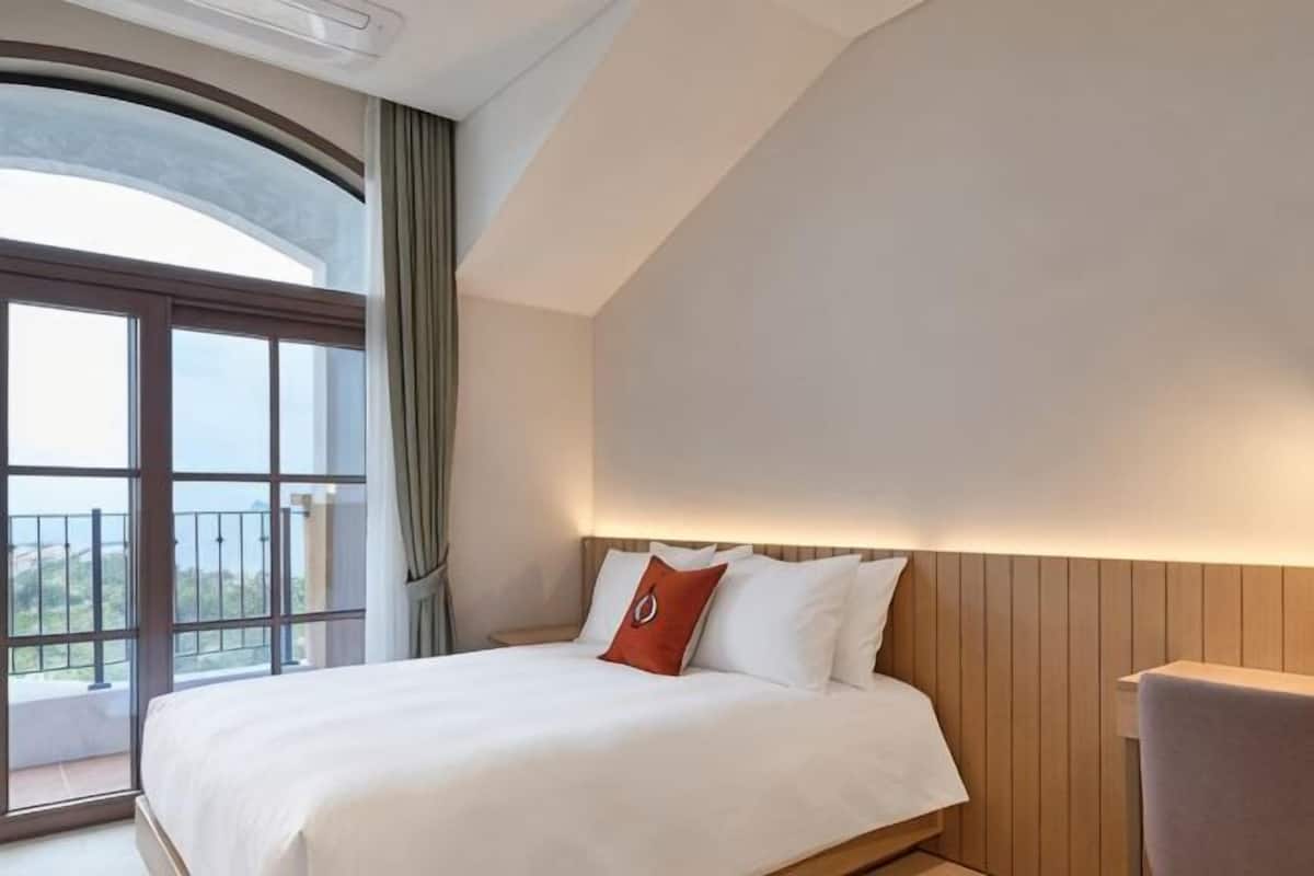 Quarto de hotel com uma cama grande, cabeceira de madeira, janela com cortinas e vista para o exterior.