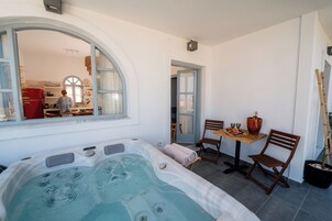 Quarto com hidromassagem, área de refeições com cadeiras e mesa, e vista para uma cozinha através de uma janela em arco.