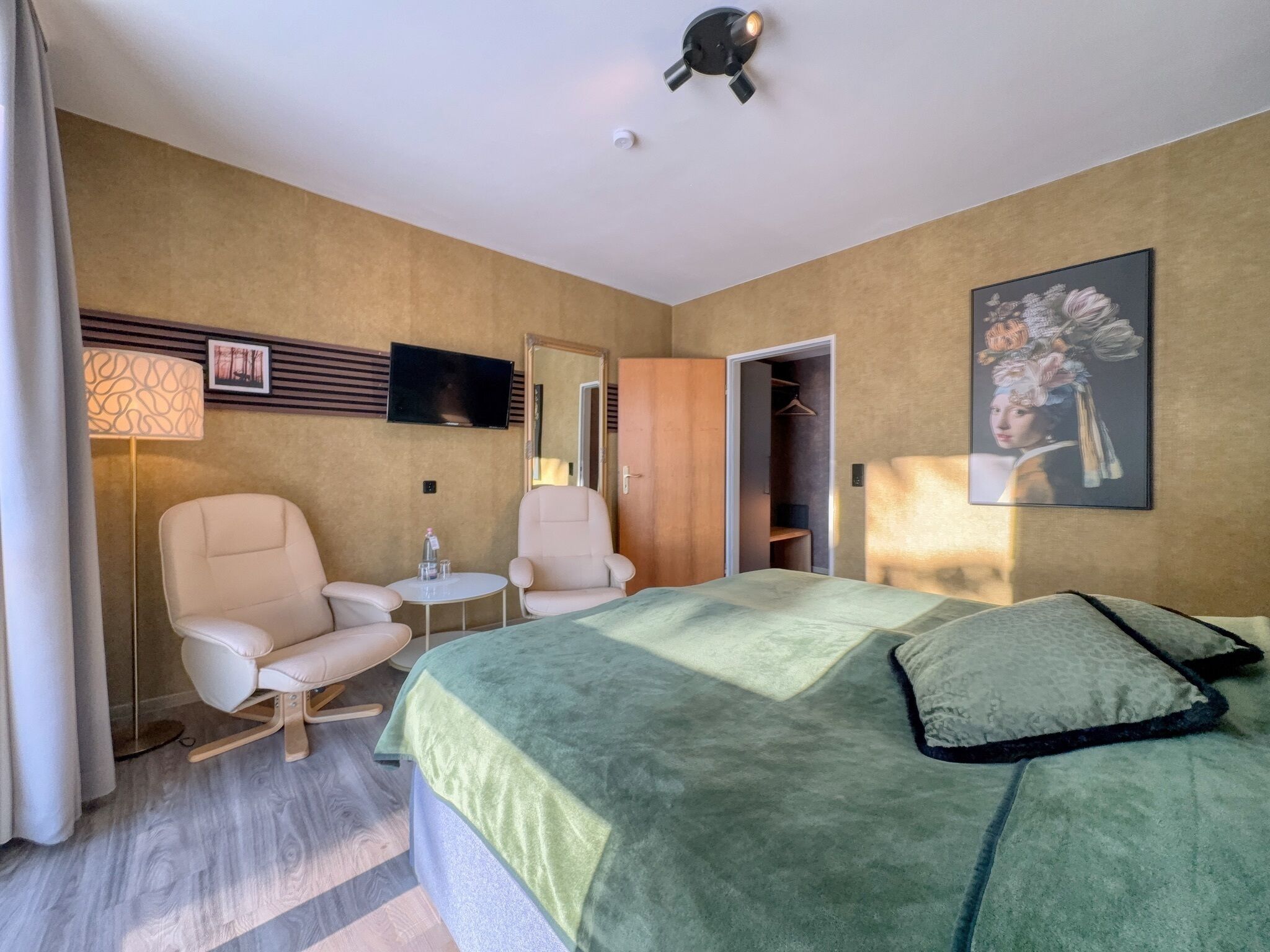 Quarto de hotel com uma cama, duas cadeiras, uma mesinha, televisão, guarda-roupa e um quadro na parede.