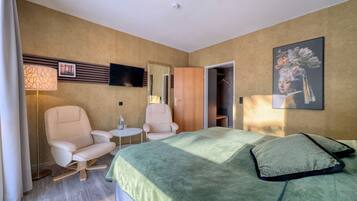 Quarto de hotel com uma cama, duas cadeiras, uma mesinha, televisão, guarda-roupa e um quadro na parede.