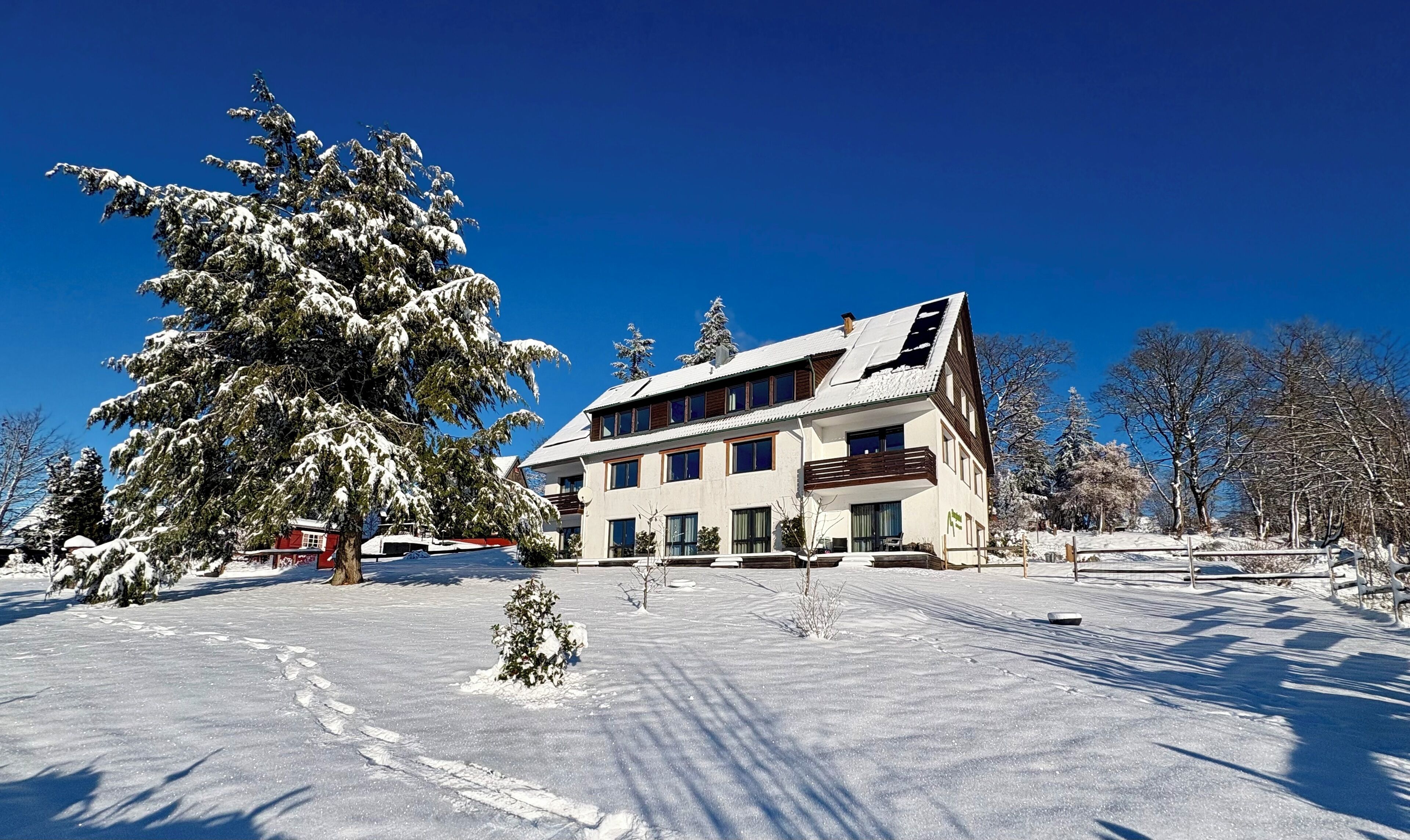 Uma casa de dois andares com varanda, rodeada por árvores cobertas de neve e um céu azul claro.
