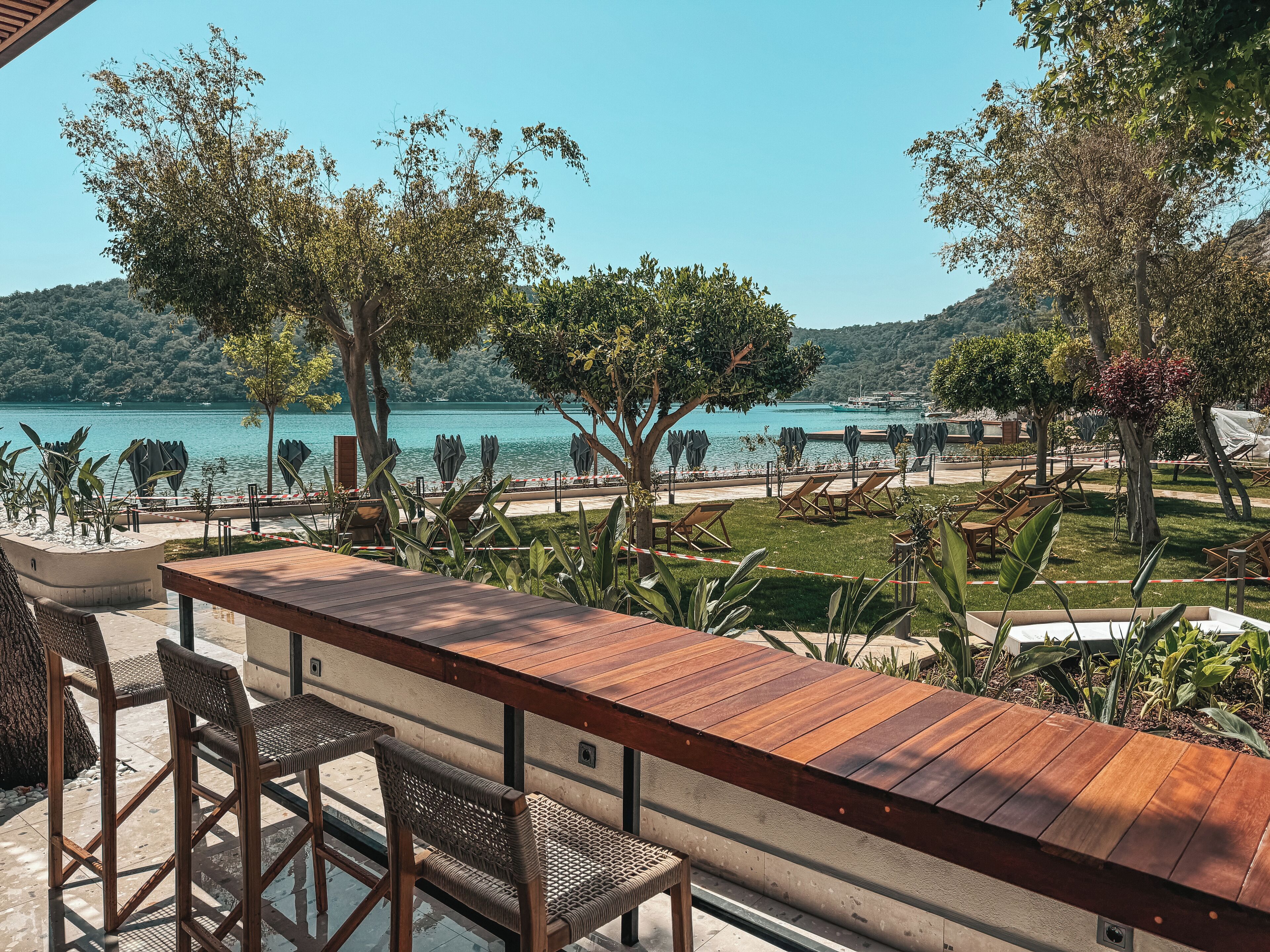 Um deck de madeira com cadeiras e uma mesa com vista para um lago, rodeado por árvores e vegetação.