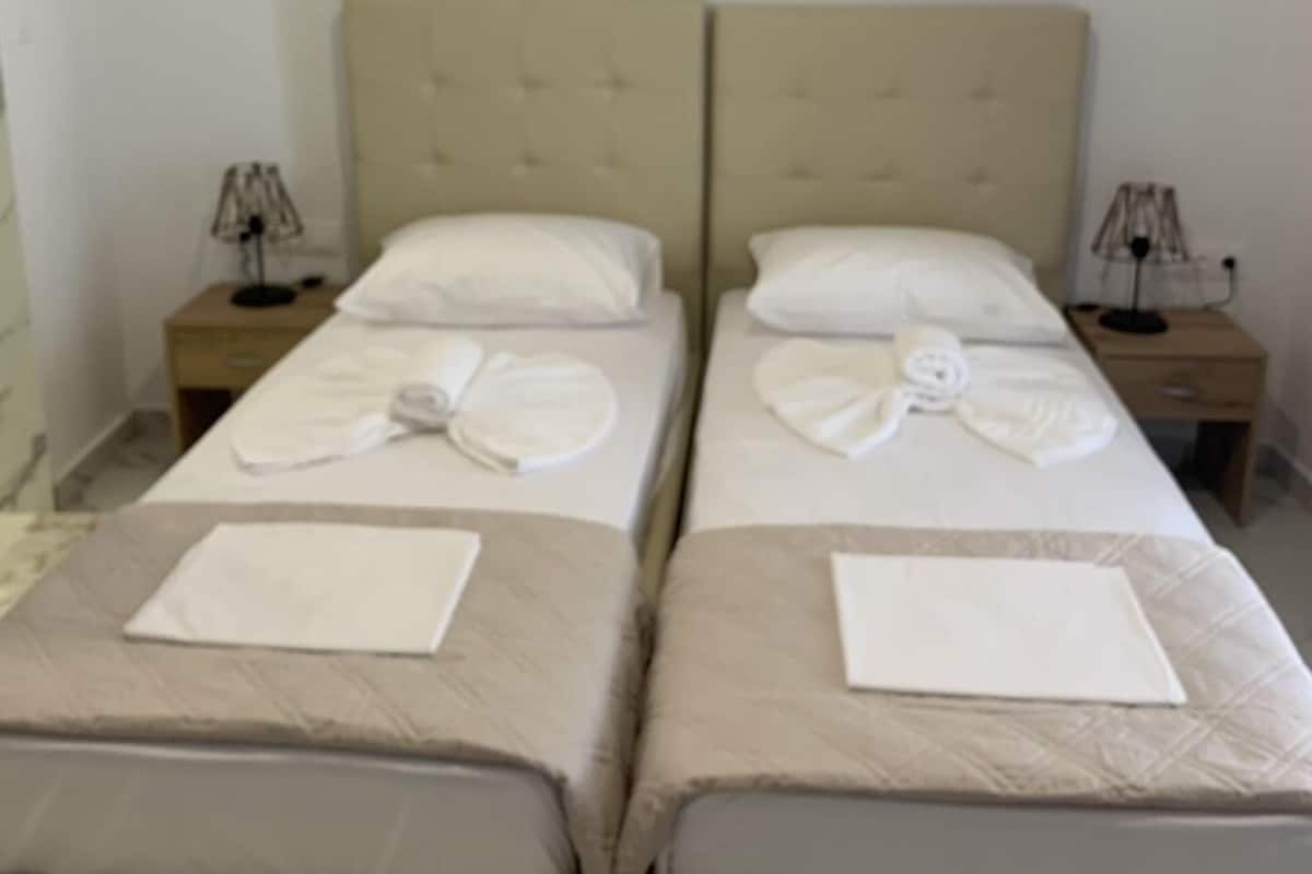 Quarto de hotel com duas camas, cada uma com cabeceira bege e roupa de cama branca. Há duas mesas de cabeceira com abajures e uma pequena mesa entre as camas.