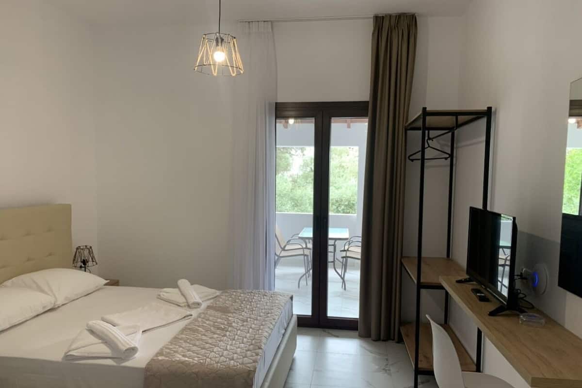 Quarto de hotel com cama, escrivaninha, televisão e varanda com cadeiras.