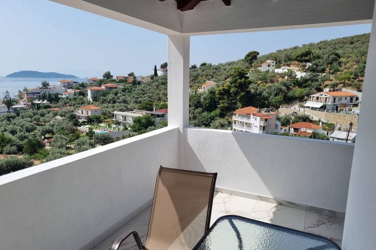Uma varanda com vista para uma vila costeira, uma cadeira e uma mesa de vidro.