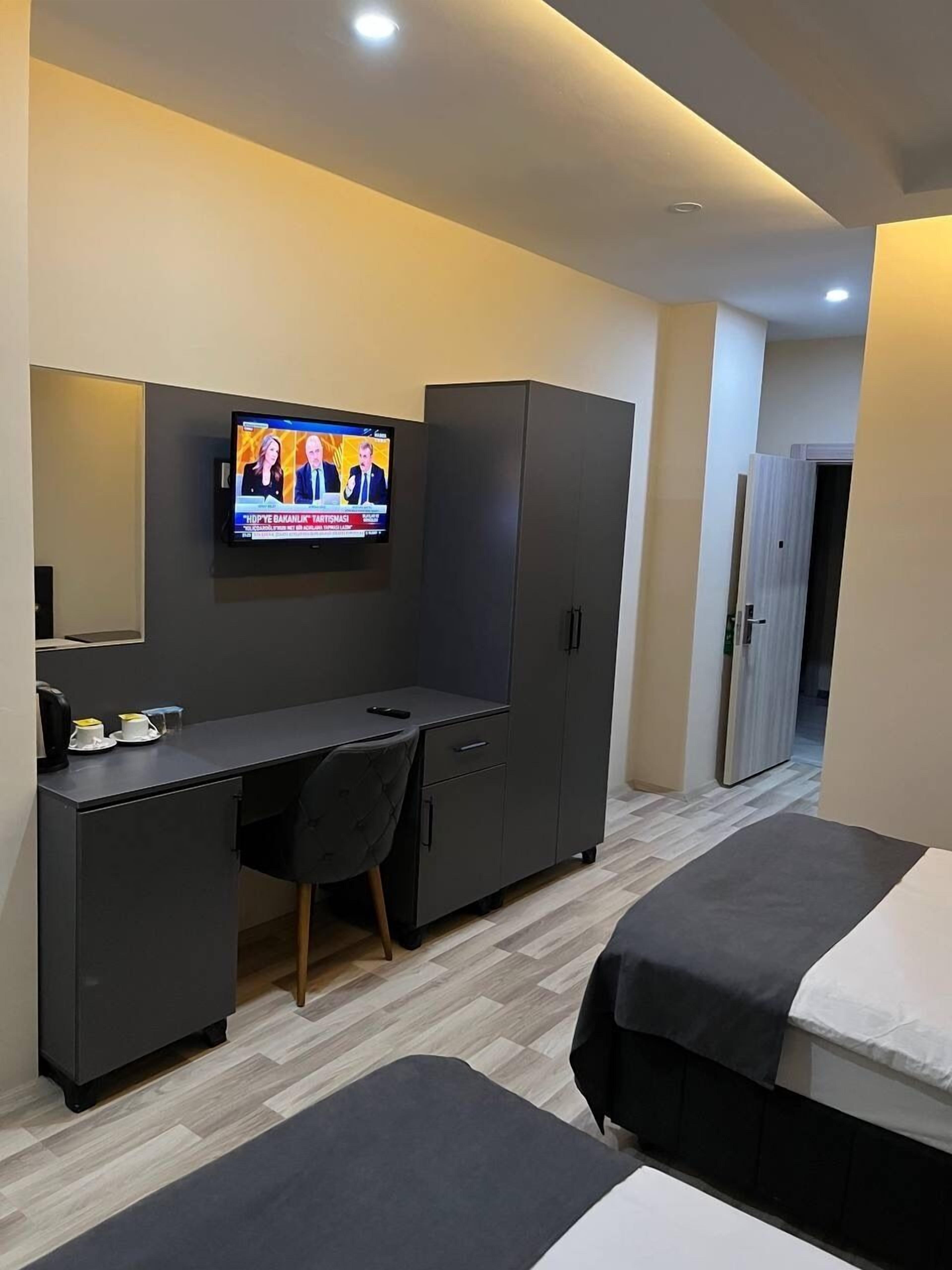 Quarto de hotel moderno com escrivaninha cinza escura, televisão na parede, cama com colcha preta e branca e um guarda-roupa.