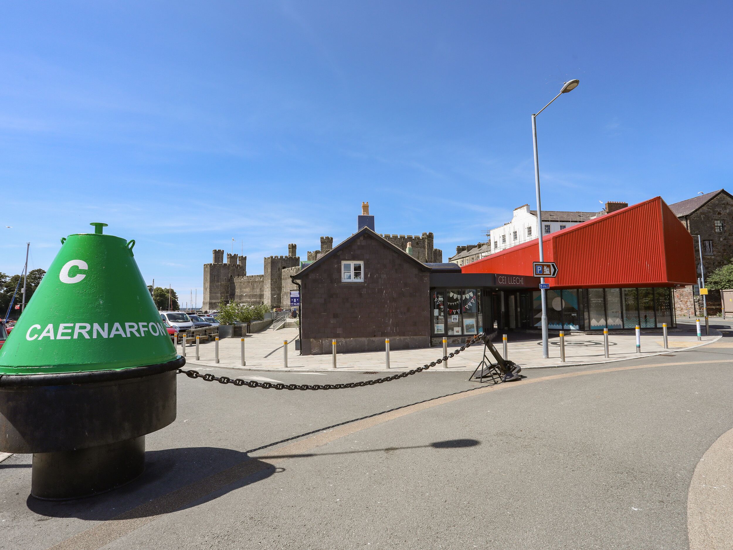 Um cone de trânsito verde com a inscrição 'CAERNARFON' numa rua, com um castelo ao fundo e um edifício de telhado vermelho com a placa 'CEI LLECHI'.