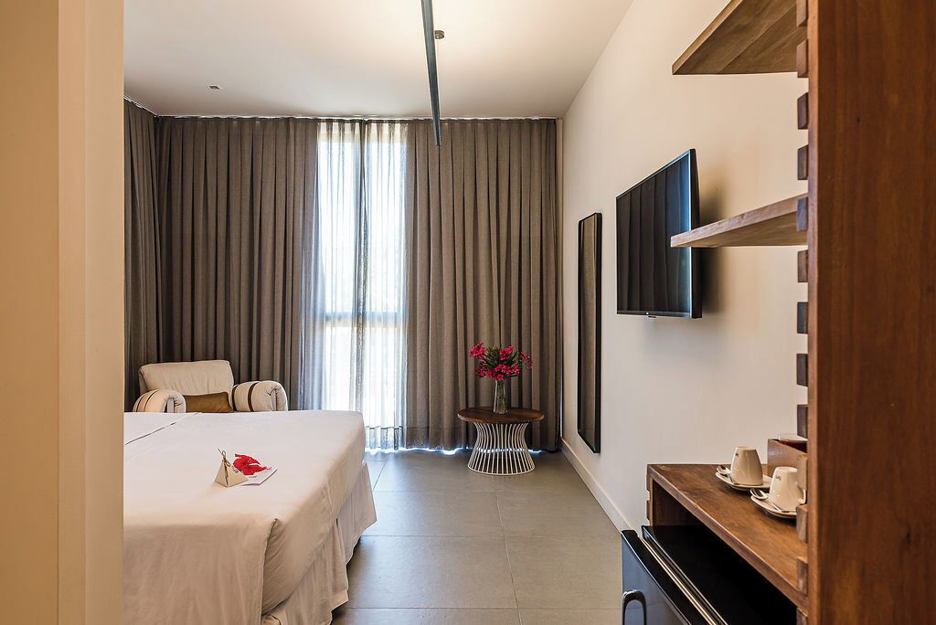 Quarto de hotel com cama, escrivaninha, cadeira, televisão e janela com cortinas.