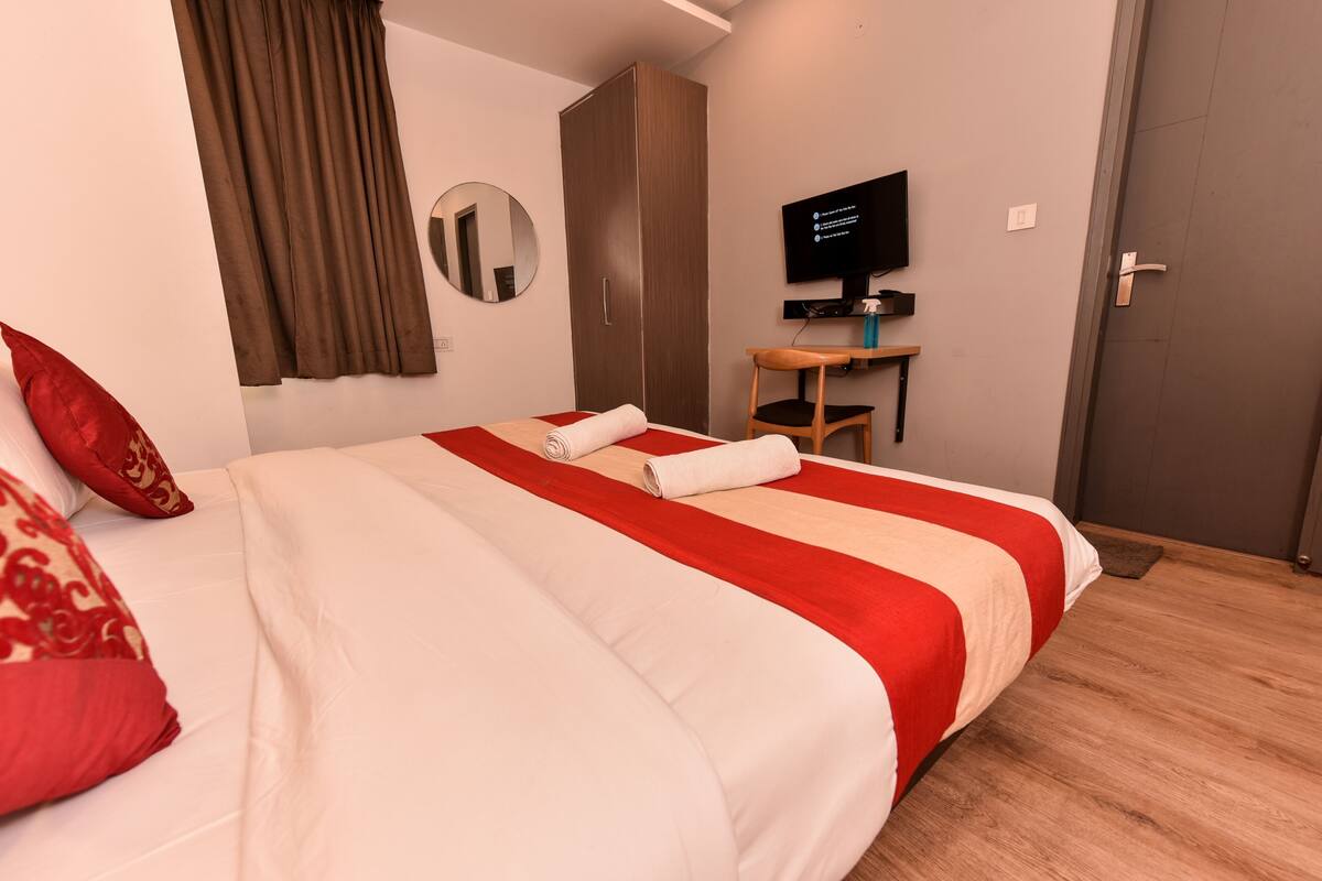 Quarto de hotel com cama, escrivaninha, televisão e guarda-roupa.