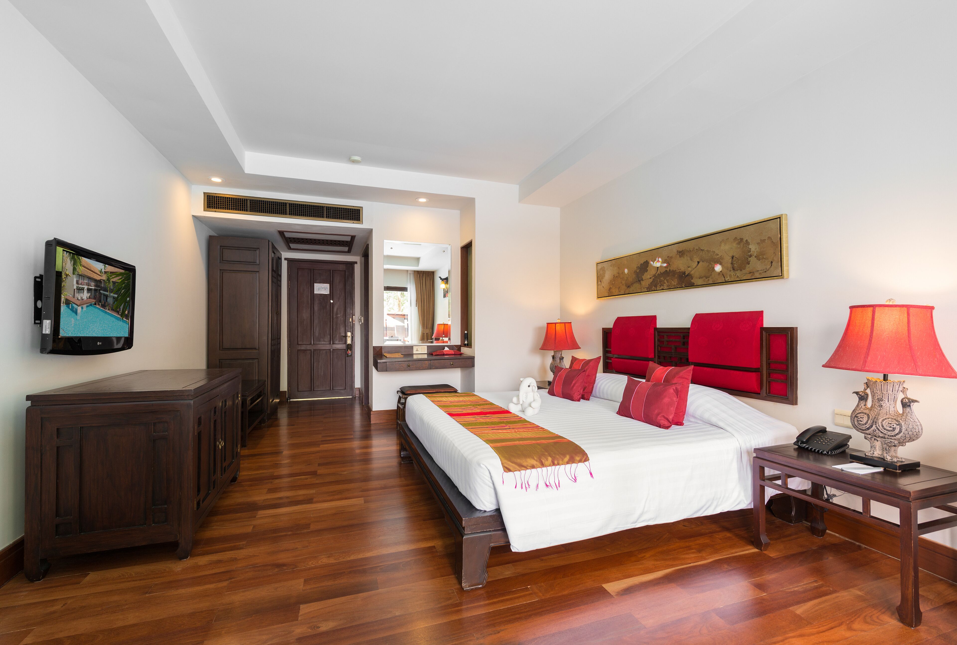 Quarto de hotel com cama grande, piso de madeira, televisão, escrivaninha e abajur vermelho.
