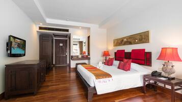 Quarto de hotel com cama grande, piso de madeira, televisão, escrivaninha e abajur vermelho.