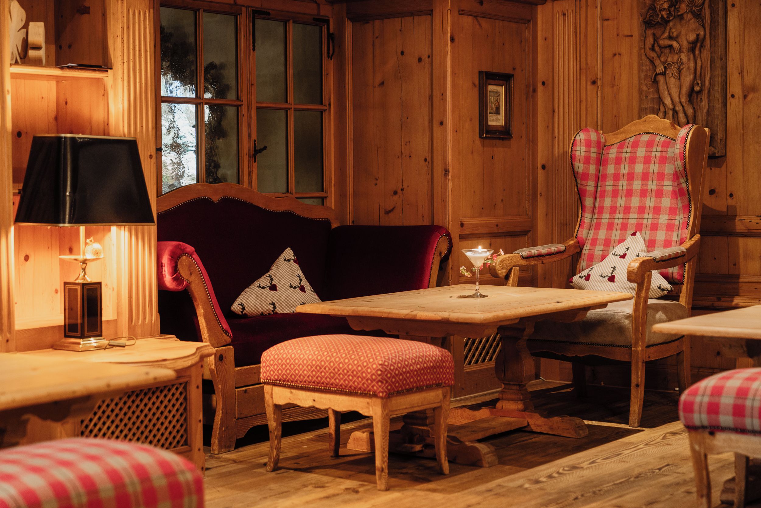 Interior acolhedor de cabana com paredes de madeira, uma poltrona de veludo vermelho, uma poltrona xadrez, uma pequena mesa de madeira e um abajur.