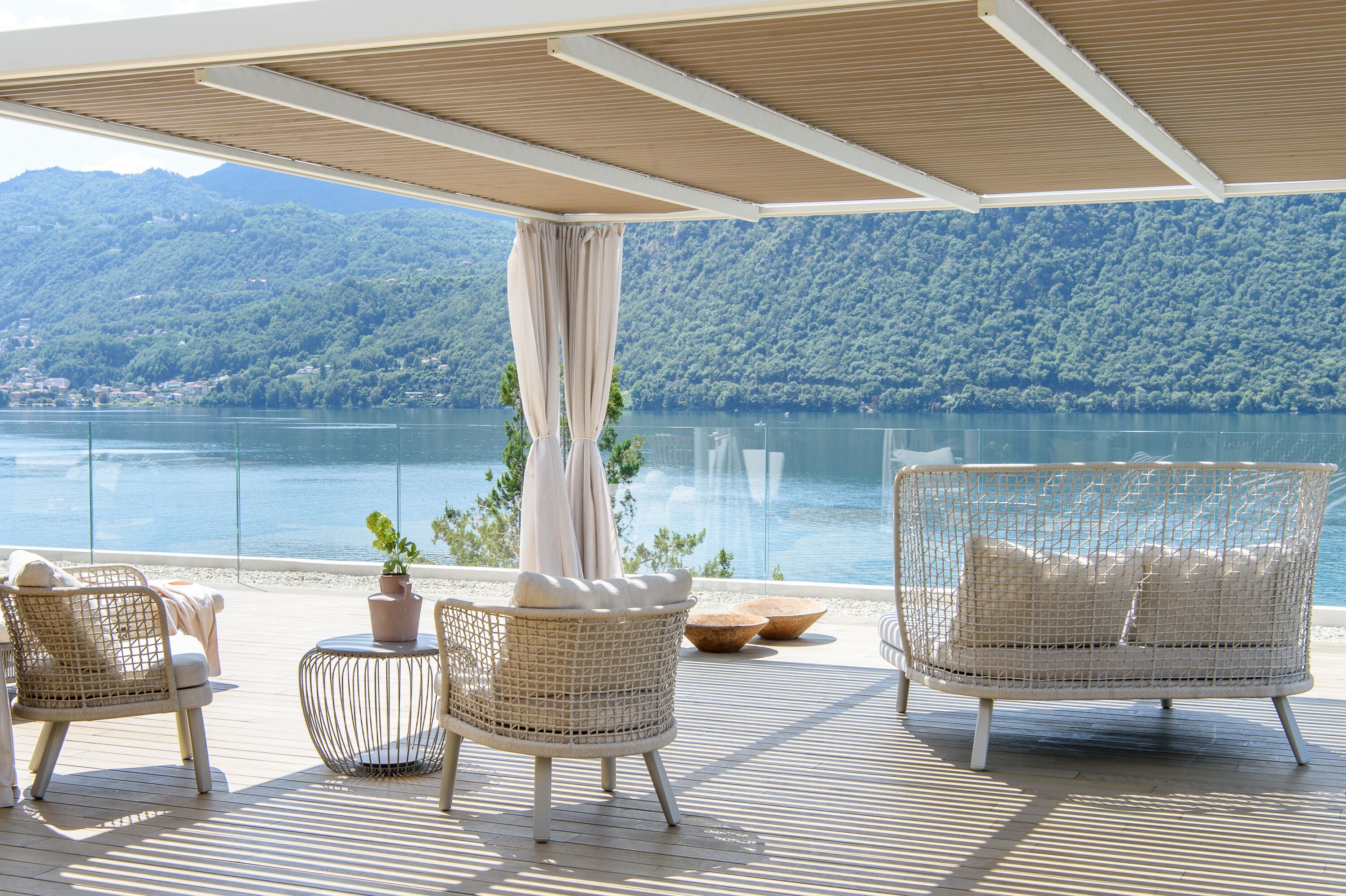 Uma área de estar coberta ao ar livre com móveis de vime, um vaso de planta e vista para um lago e montanhas.