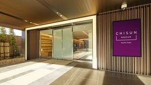 Uma entrada de prédio moderna com porta de vidro, uma placa com a inscrição "CHISUN PREMIUM Kyoto Kujo" e teto estampado.