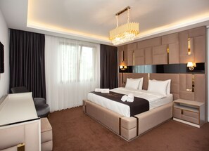 Quarto de hotel moderno com uma cama grande, uma escrivaninha e uma cadeira. Há um candelabro sobre a cama e luminárias fixadas na parede em ambos os lados.