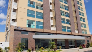 Um hotel de vários andares com design moderno, destacando uma entrada imponente e uma área ajardinada com plantas pequenas e uma palmeira.