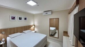 Quarto de hotel com cama, mesa de cabeceira, abajur, espelho e televisão.
