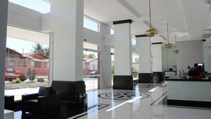 Um saguão de hotel com balcão de recepção, cadeiras de couro preto, piso ladrilhado com padrão geométrico e grandes janelas com vista para o exterior.