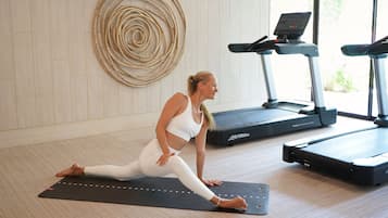 Mulher fazendo uma pose de yoga em um tapete, num espaço com uma esteira e um objeto circular fixado na parede.