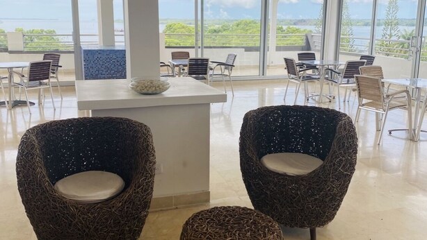 Amplo espaço interno com grandes janelas que oferecem vista para o mar. Há várias cadeiras e uma pequena mesa com uma tigela decorativa.