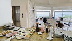 Um buffet de café da manhã com diversos alimentos e vista para o mar através de grandes janelas.