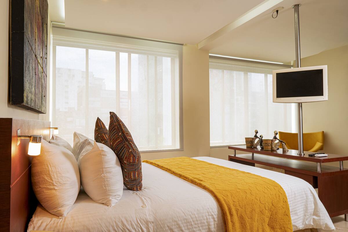 Quarto de hotel com uma cama grande, uma escrivaninha com televisão de tela plana e uma janela com persianas.