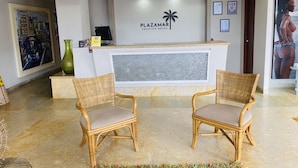 Uma área de recepção com uma placa indicando 'Plazamar Pacifico Hotel'. Há duas cadeiras de vime em frente ao balcão da recepção.