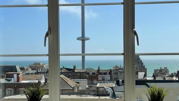 Janela com vista para um horizonte urbano e o mar, exibindo uma torre alta e duas plantas em vasos no parapeito.