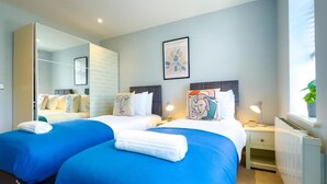 Quarto de hotel com duas camas, um espelho grande, uma obra de arte emoldurada, uma mesa de cabeceira com abajur e uma janela com planta.
